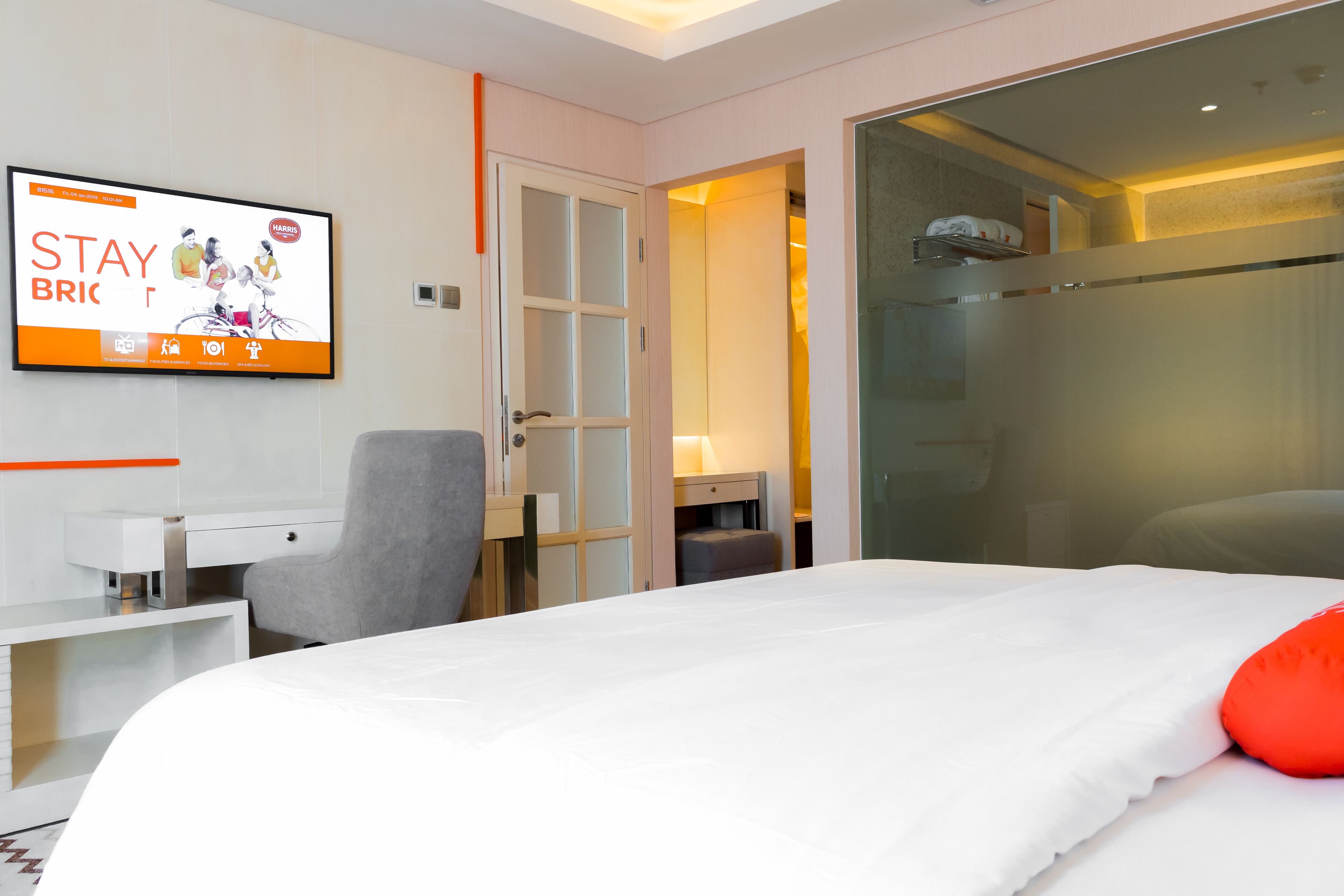 Quarto de hotel com cama, televisão exibindo um evento de ciclismo, poltrona cinza e box de chuveiro de vidro.