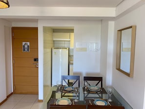 Uma sala de jantar com uma mesa de vidro, cadeiras e uma cozinha ao fundo.