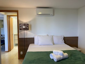 Quarto de hotel com cama, manta verde, almofadas brancas, abajur e ar condicionado.