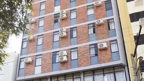 Um edifício de vários andares com design moderno, caracterizado por janelas amplas e aparelhos de ar condicionado. No andar térreo, há lojas, incluindo uma loja de moda e um hotel chamado 'Hotel Romanville'.