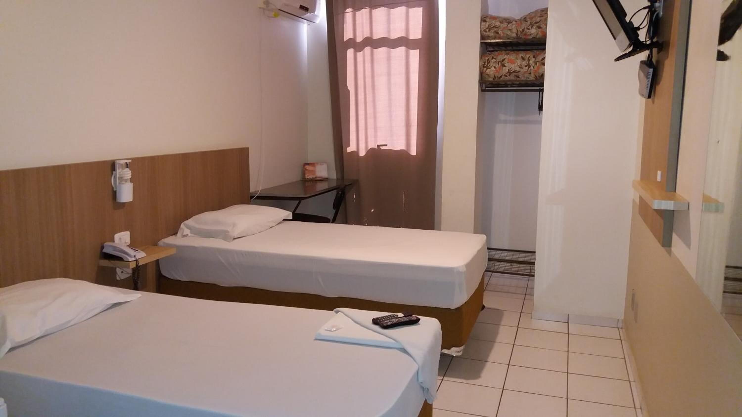Quarto de hotel pequeno e compacto, com duas camas de solteiro, uma mesinha de cabeceira com telefone e uma pequena escrivaninha com luminária.