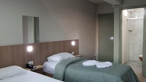 Quarto de hotel com duas camas de solteiro, uma mesinha de cabeceira com telefone, e um banheiro visível através de uma porta aberta.