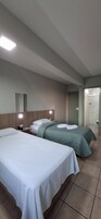 Quarto de hotel com duas camas de solteiro, uma mesinha de cabeceira com telefone, e um banheiro visível através de uma porta aberta.