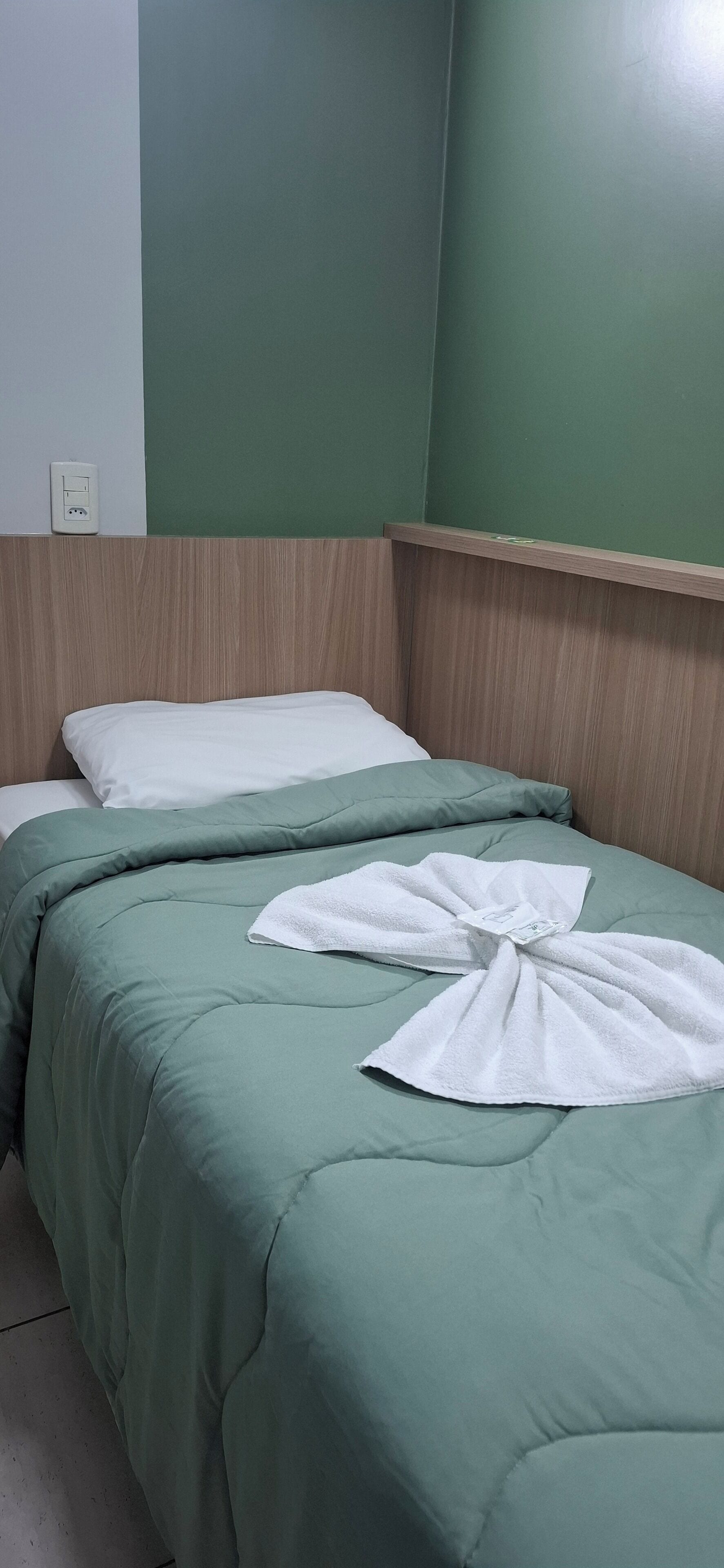 Cama de casal com colcha verde e almofada branca. Uma toalha branca dobrada está no centro da cama.