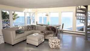 Uma sala de estar ampla com um sofá seccional grande, um banquinho e vista para o oceano através de amplas janelas.