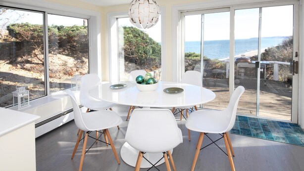 Uma área de refeições moderna com uma mesa e cadeiras brancas, grandes janelas com vista para o mar e uma luminária decorativa em formato de globo.