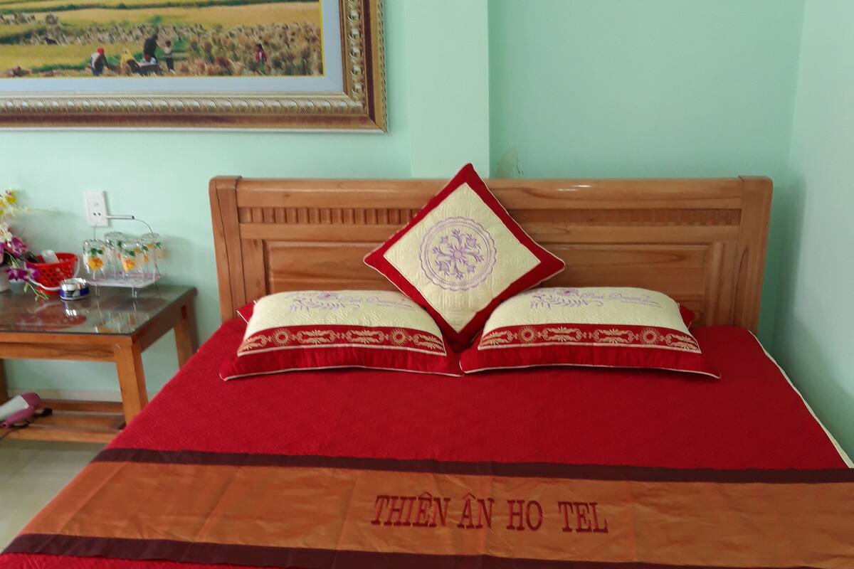 Cama com edredom vermelho e castanho, duas almofadas e uma toalha branca dobrada com cuidado.