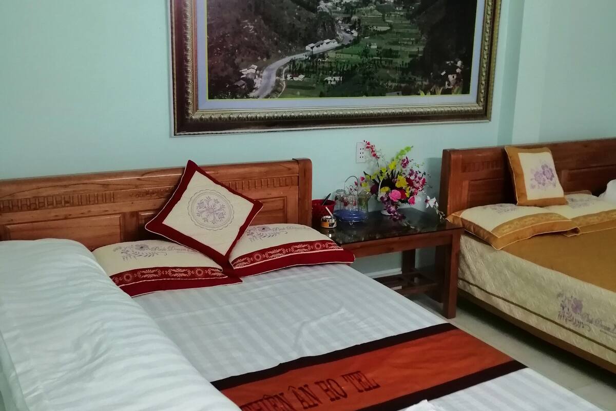 Quarto de hotel com duas camas, um quadro de paisagem emoldurado, um vaso com flores e uma cabeceira de madeira.