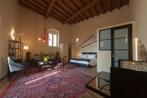 Quarto aconchegante com cama, sofá, mesa de centro e tapete. Há uma janela com cortinas, um candelabro e uma escada.