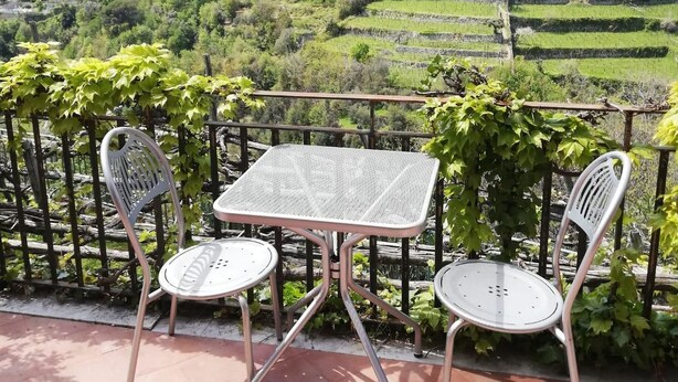 Um terraço com mesa e duas cadeiras, com vista para uma aldeia em socalcos com edifícios e vegetação.