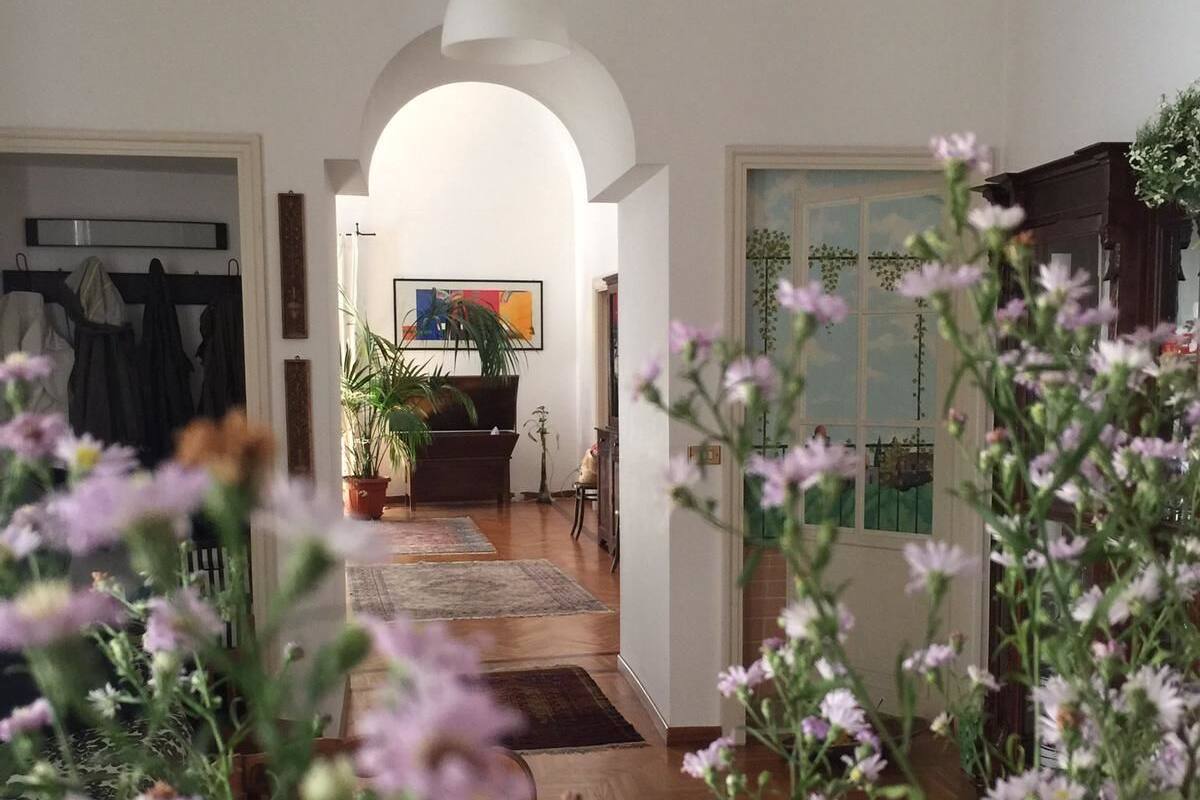 Um interior moderno com um arco central que dá para uma sala de estar, uma mesa de jantar e um sofá. Há plantas em vasos e um vaso com flores em primeiro plano.