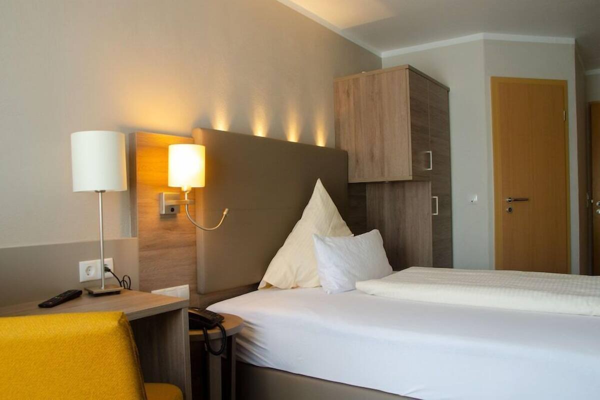 Quarto de hotel com cama, mesa de cabeceira, luminária e cabeceira de madeira.
