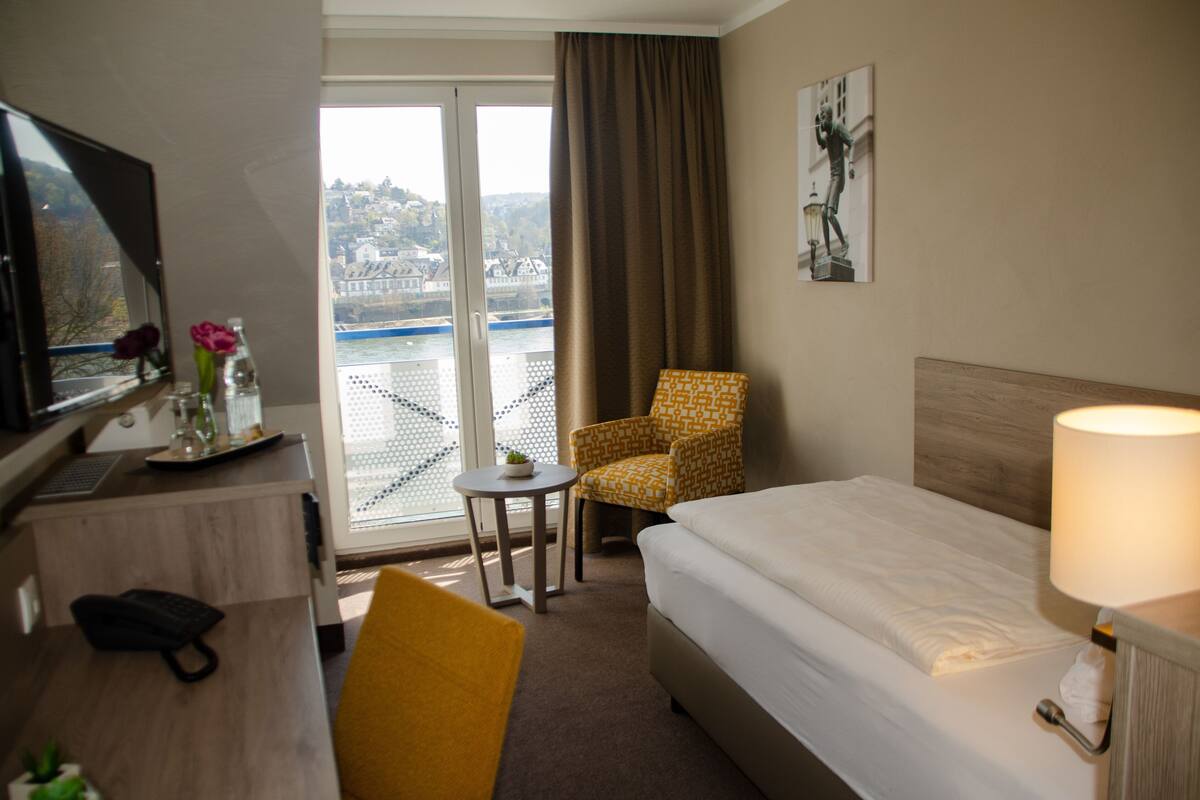 Quarto de hotel com uma cama, uma escrivaninha com um computador, uma cadeira, uma mesinha com uma planta e vista para um corpo de água através de uma janela.