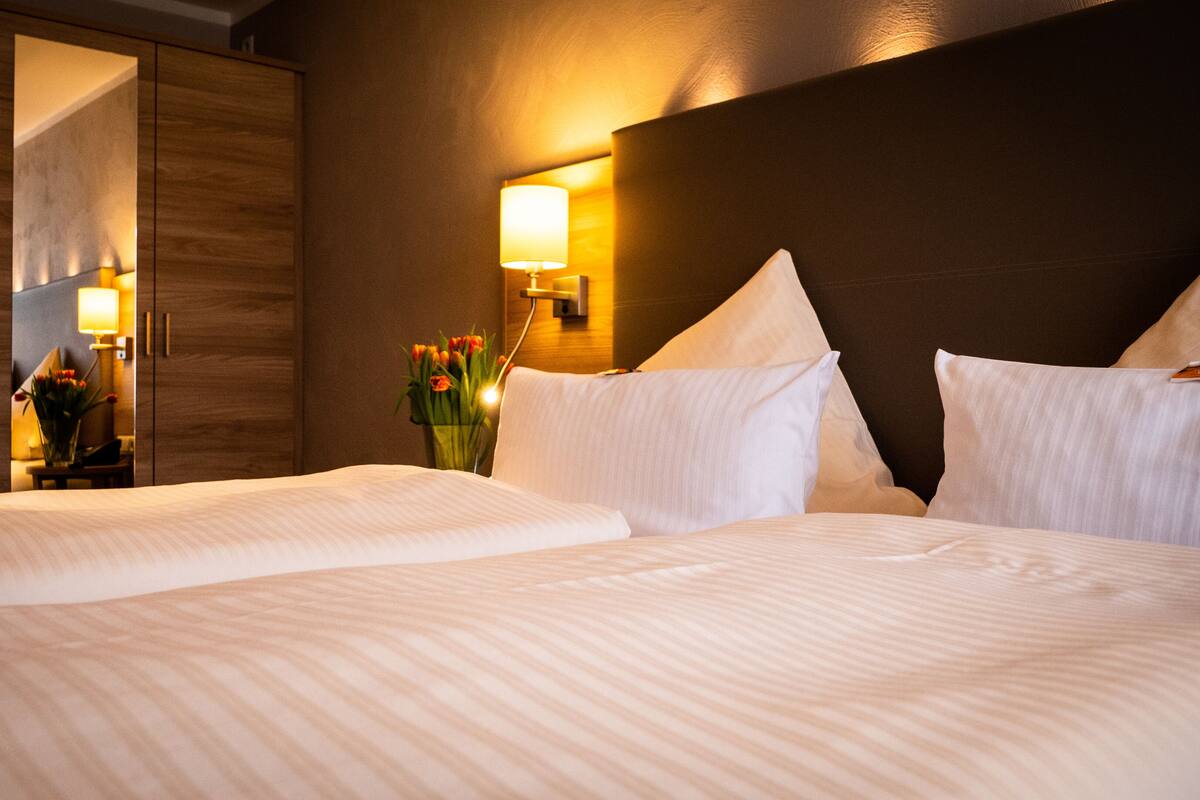 Quarto de hotel com uma cama grande, um guarda-roupa, uma mesa de cabeceira com abajur e um vaso com flores.