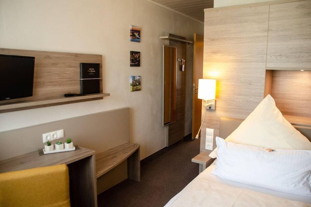 Quarto de hotel com uma cama, uma escrivaninha com cadeira, televisão, prateleira na parede e um candeeiro de parede.