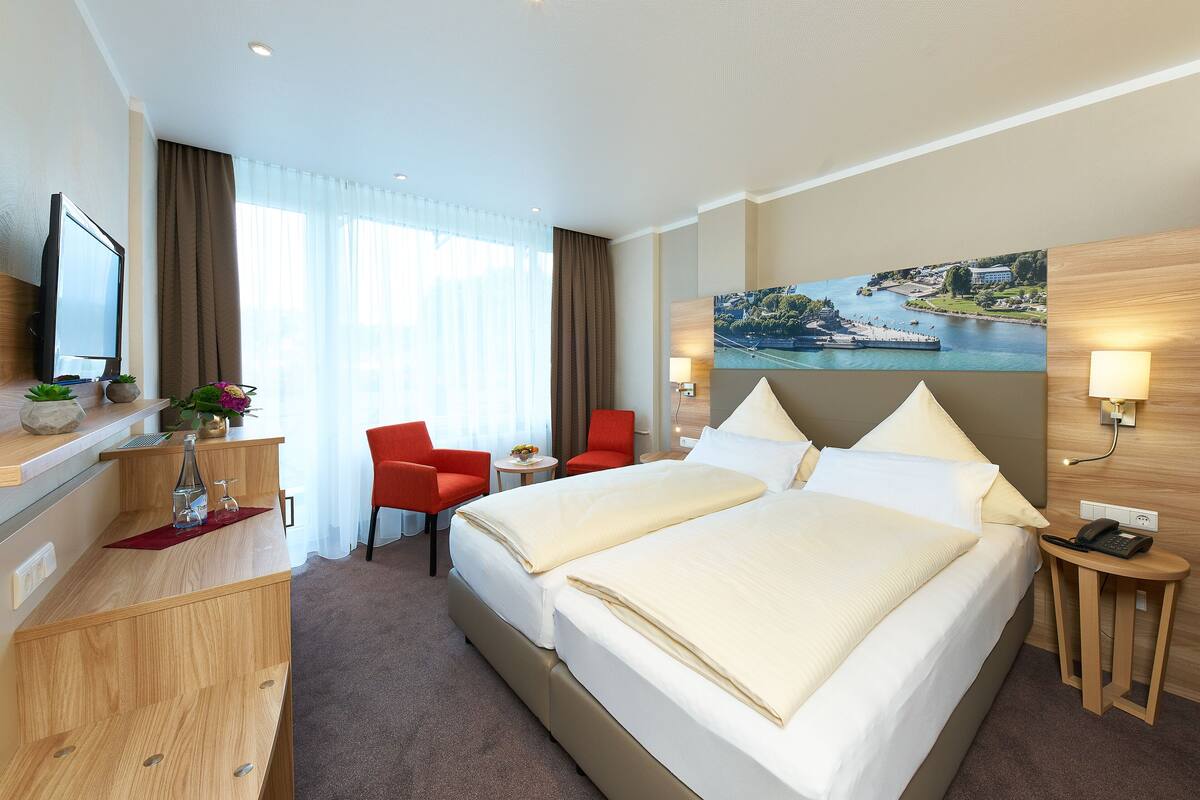 Quarto de hotel com uma cama grande, uma escrivaninha com televisão, uma cadeira e uma mesinha de cabeceira com abajur.