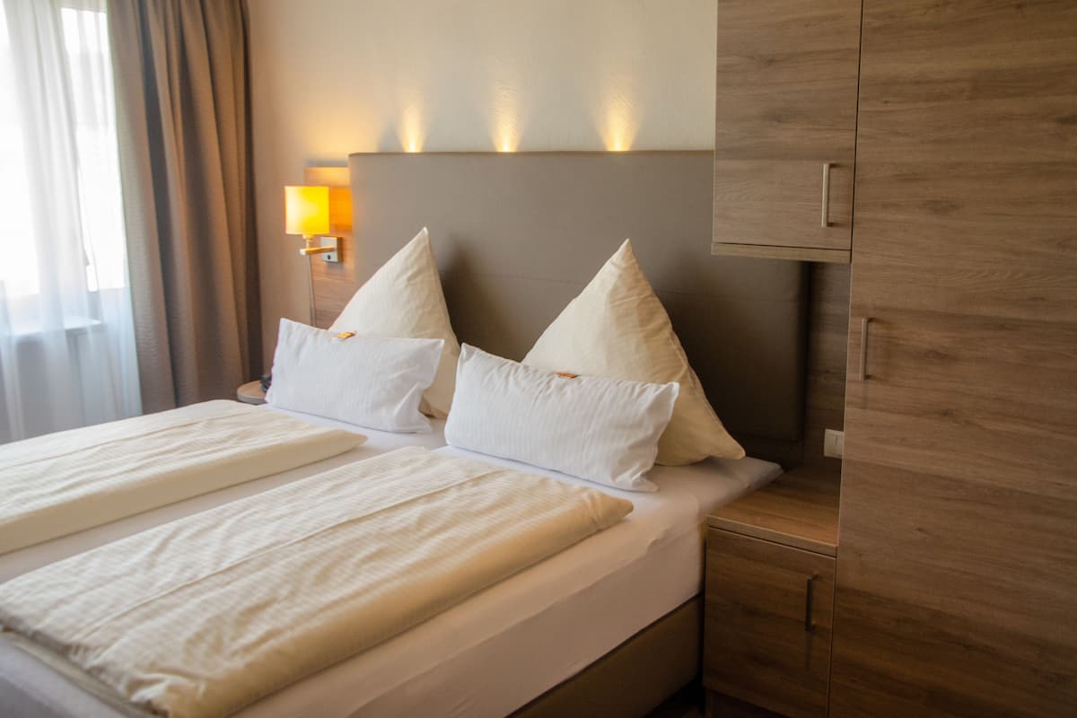 Quarto de hotel com uma cama grande, duas almofadas, um abajur e um guarda-roupa de madeira.