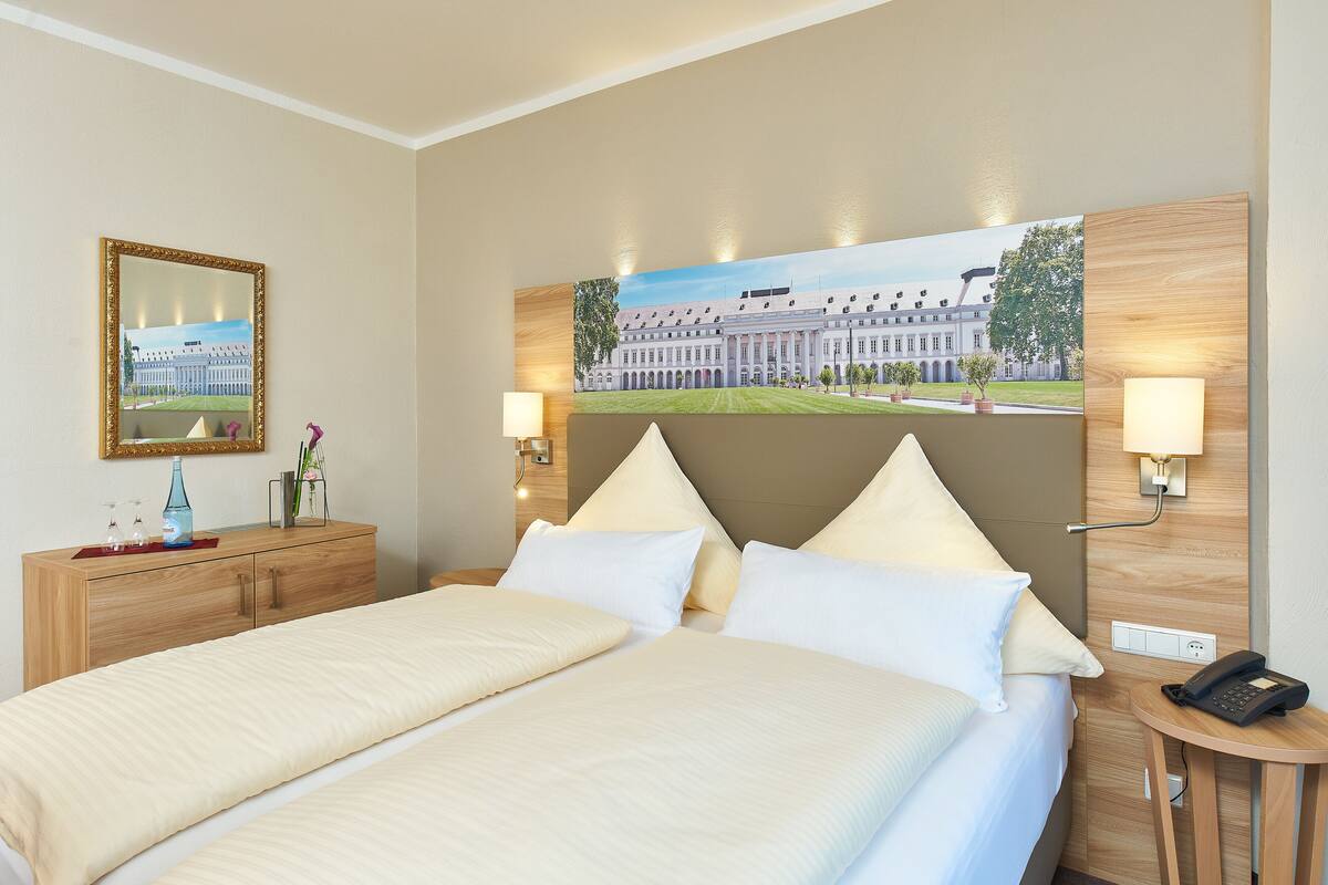 Quarto de hotel com uma cama grande, penteadeira de madeira, um espelho e um quadro de um edifício na parede.