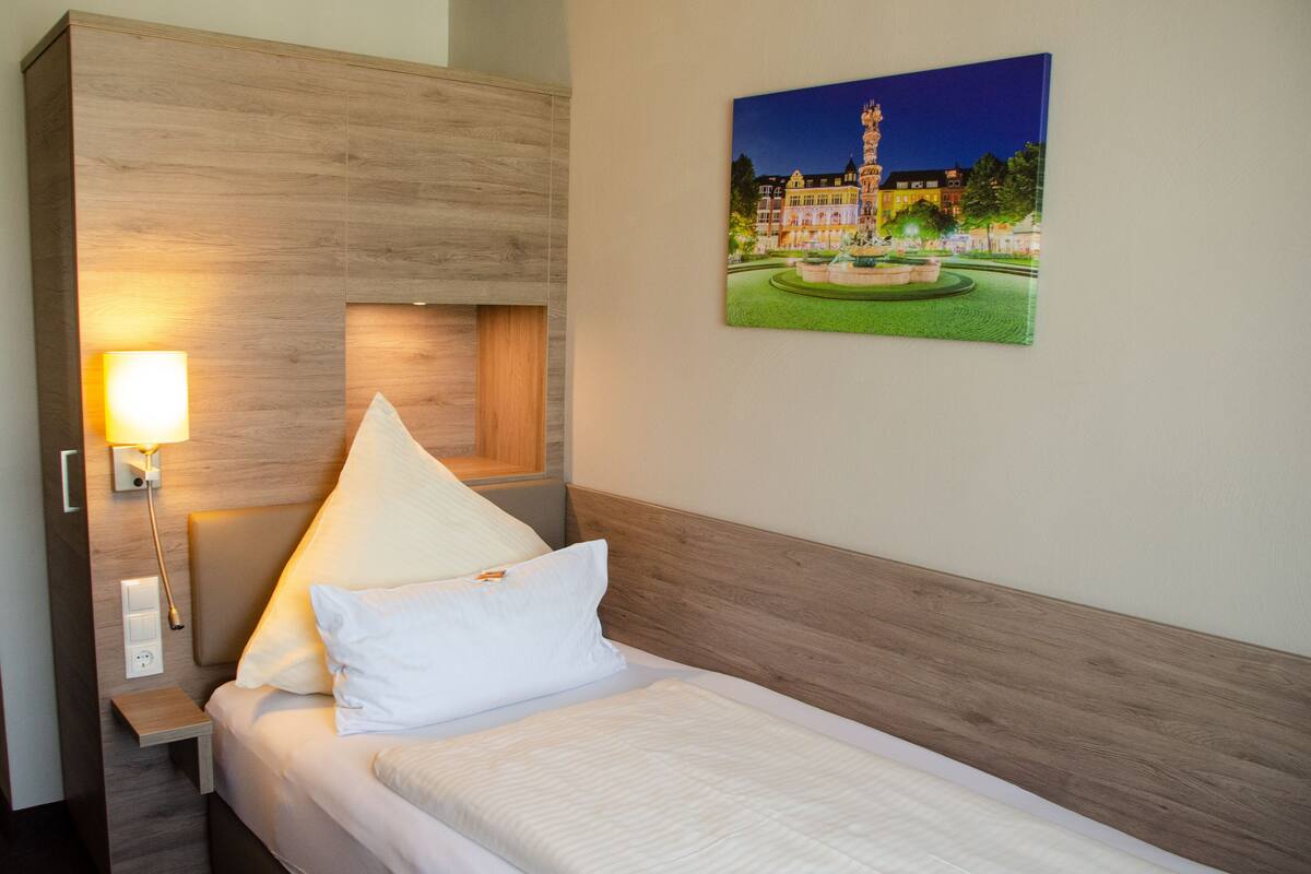 Quarto de hotel com uma cama, uma mesa de cabeceira com abajur, cabeceira de madeira e uma pintura de uma paisagem urbana na parede.