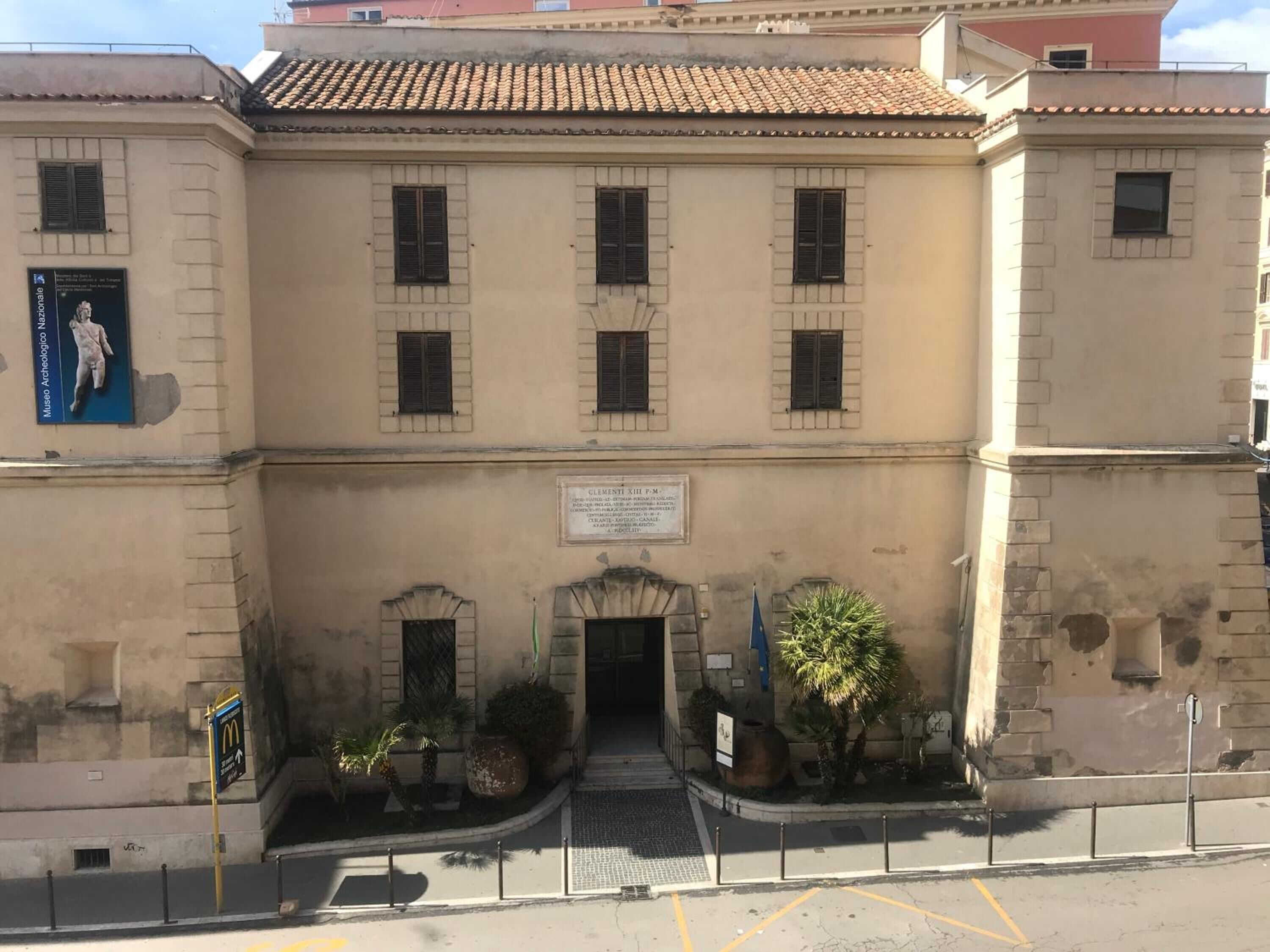 Um edifício histórico com telhado em azulejos, entrada central e placa sobre a porta.