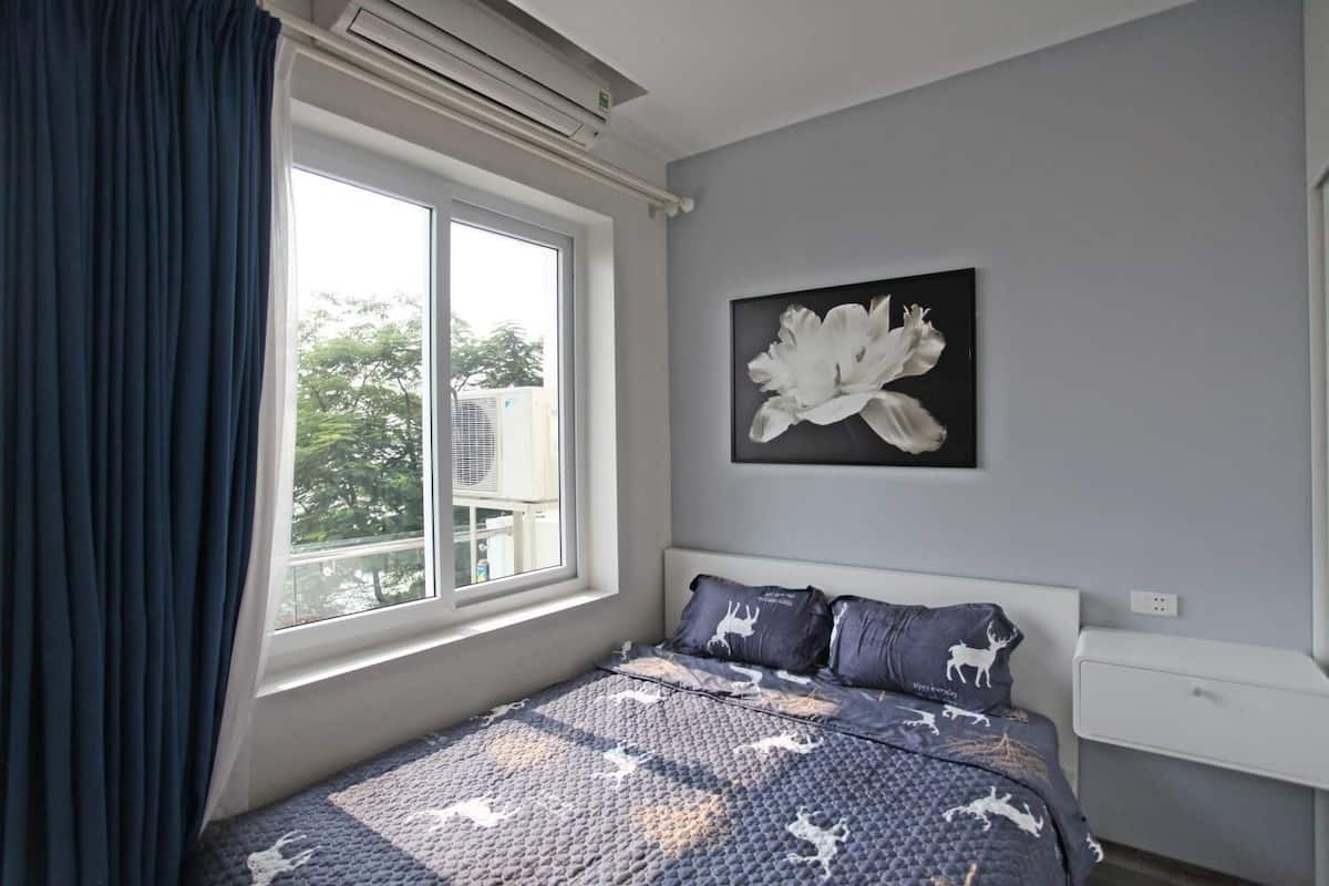 Quarto com uma janela ampla, cama com roupa de cama estampada, ar condicionado fixado na parede e uma obra de arte emoldurada.