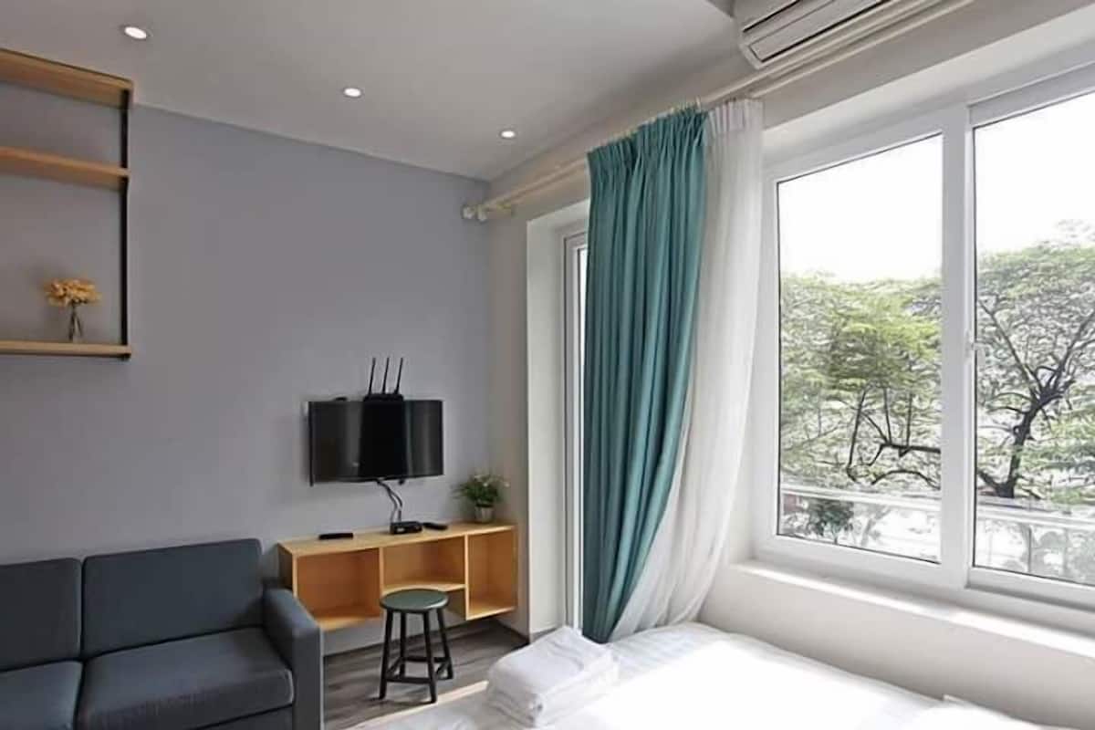 Quarto de hotel moderno com cama, sofá, televisão na parede e janela com cortinas.