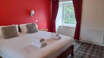 Quarto de hotel com cama, paredes vermelhas, janela com cortinas, mesa de cabeceira e abajur.