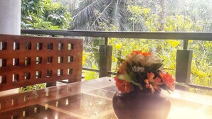 Uma mesa de madeira com tampo de vidro, um banco e um vaso de flores numa varanda com vista para a vegetação tropical.