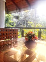 Uma mesa de madeira com tampo de vidro, um banco e um vaso de flores numa varanda com vista para a vegetação tropical.