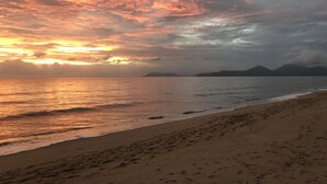 Uma praia com pegadas que levam à água, um mar calmo e montanhas distantes sob um céu nublado.