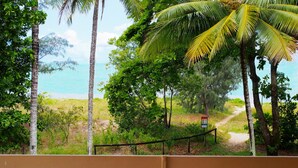 Vista de uma varanda com mesa e cadeiras, para uma praia com palmeiras e um caminho que leva a uma placa.