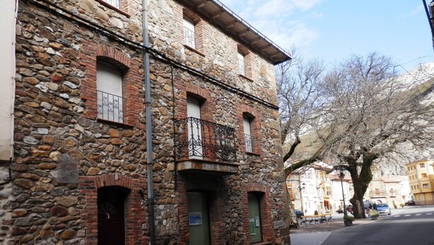 Um edifício de pedra com várias janelas e uma varanda, situado numa rua com calçada e árvores.