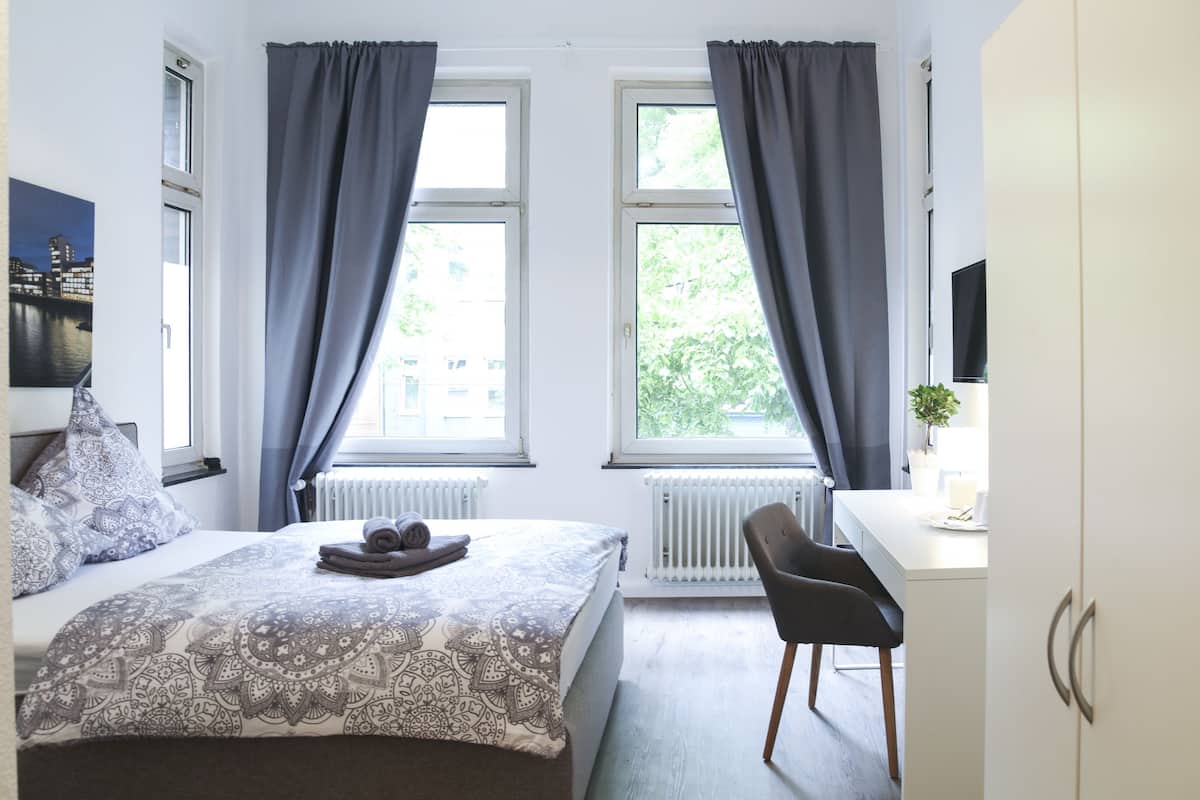 Quarto com cama, escrivaninha e cadeira. Há duas janelas com cortinas, um radiador e uma planta na escrivaninha.
