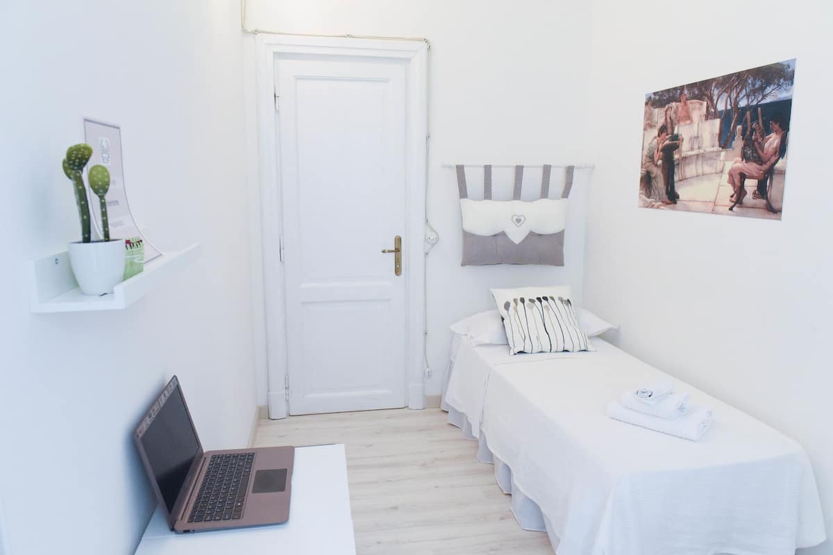 Um quarto pequeno e bem arrumado, com uma cama de solteiro, uma mesinha de cabeceira, um laptop e uma prateleira na parede com um cacto.