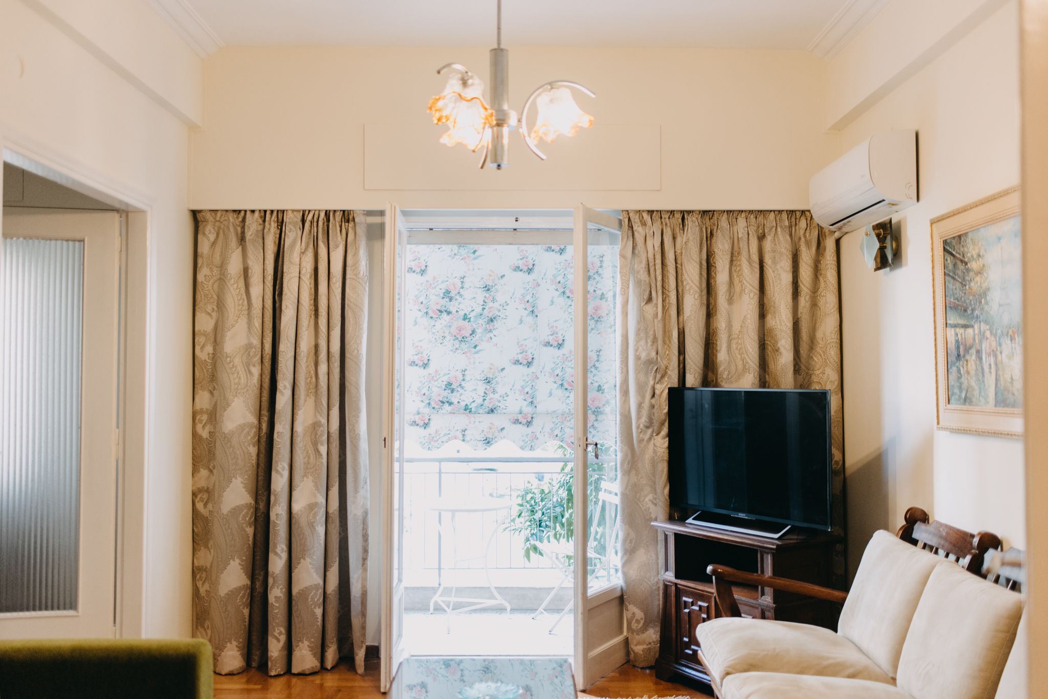Uma sala de estar com televisão, sofá, lustre e varanda com cortinas.