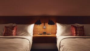 Quarto de hotel com duas camas, uma mesinha de cabeceira e duas luminárias fixadas na parede.