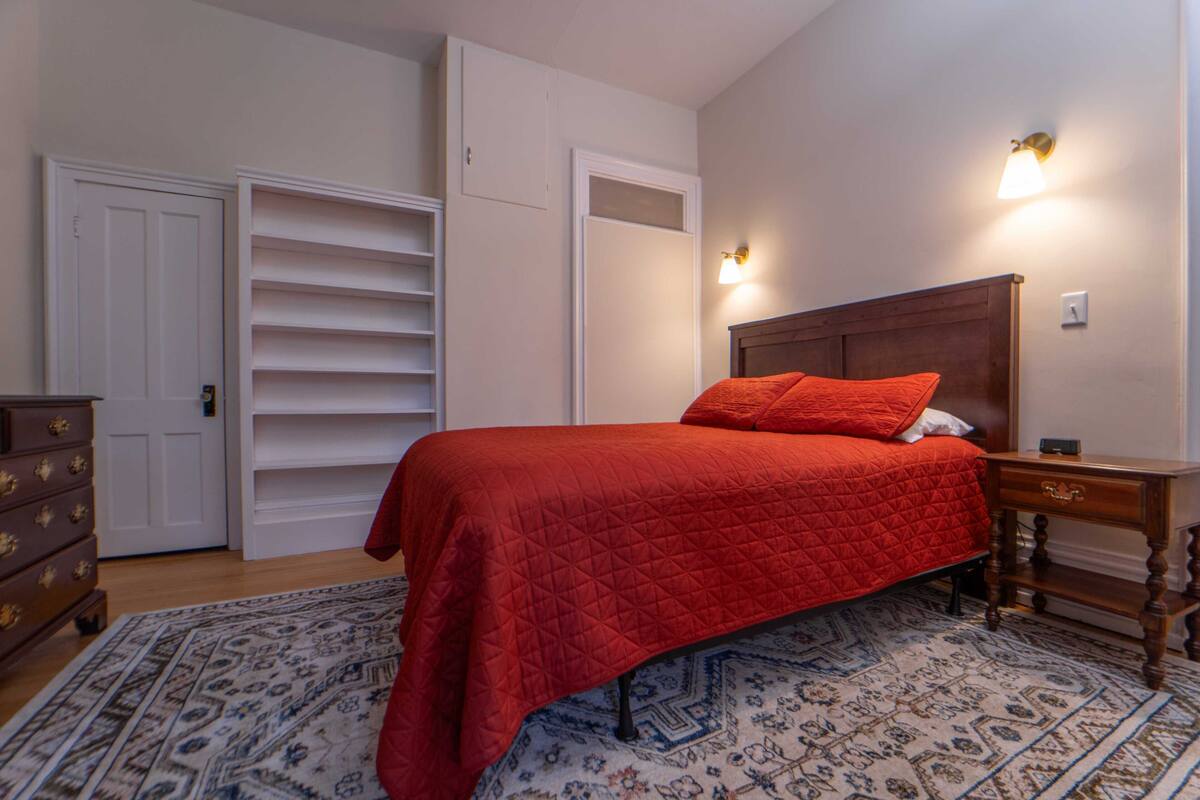 Quarto com colcha vermelha, mesa de cabeceira de madeira, tapete estampado e armários embutidos.