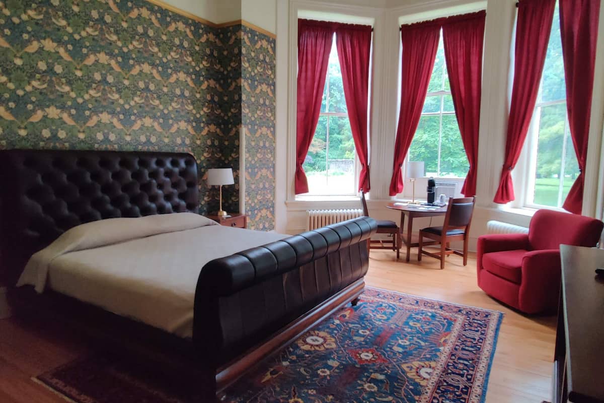 Quarto com uma cama grande, uma cadeira vermelha, uma mesa pequena e vista para o verde lá fora.