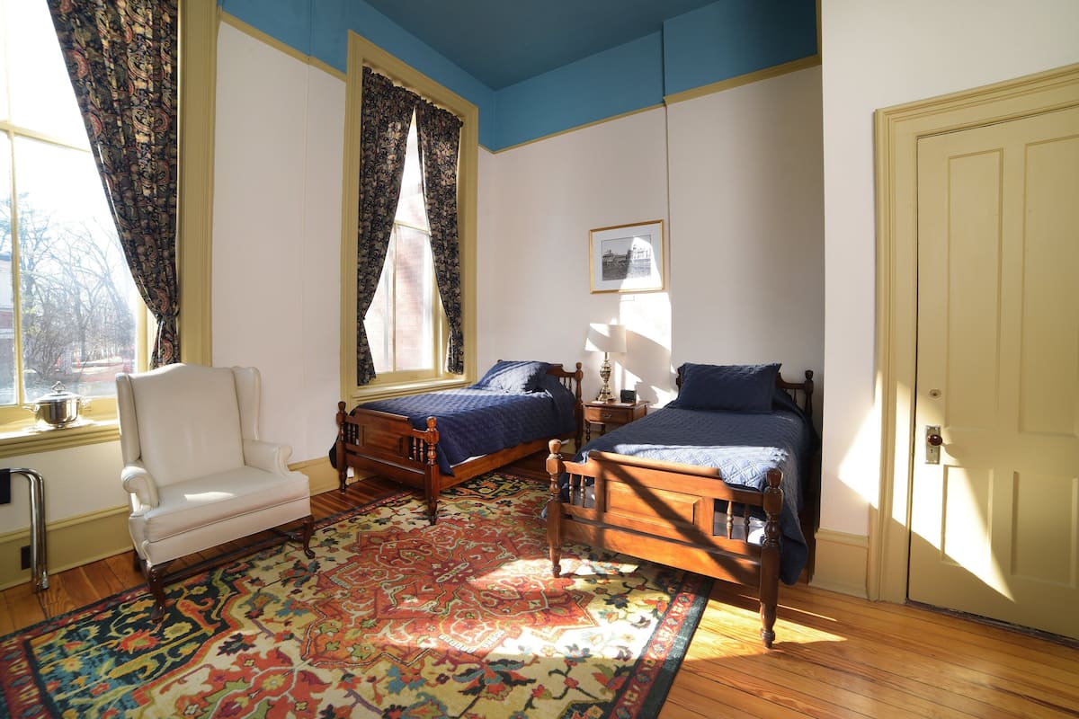 Quarto com duas camas, uma cadeira, janela com cortinas, tapete e um quadro na parede.