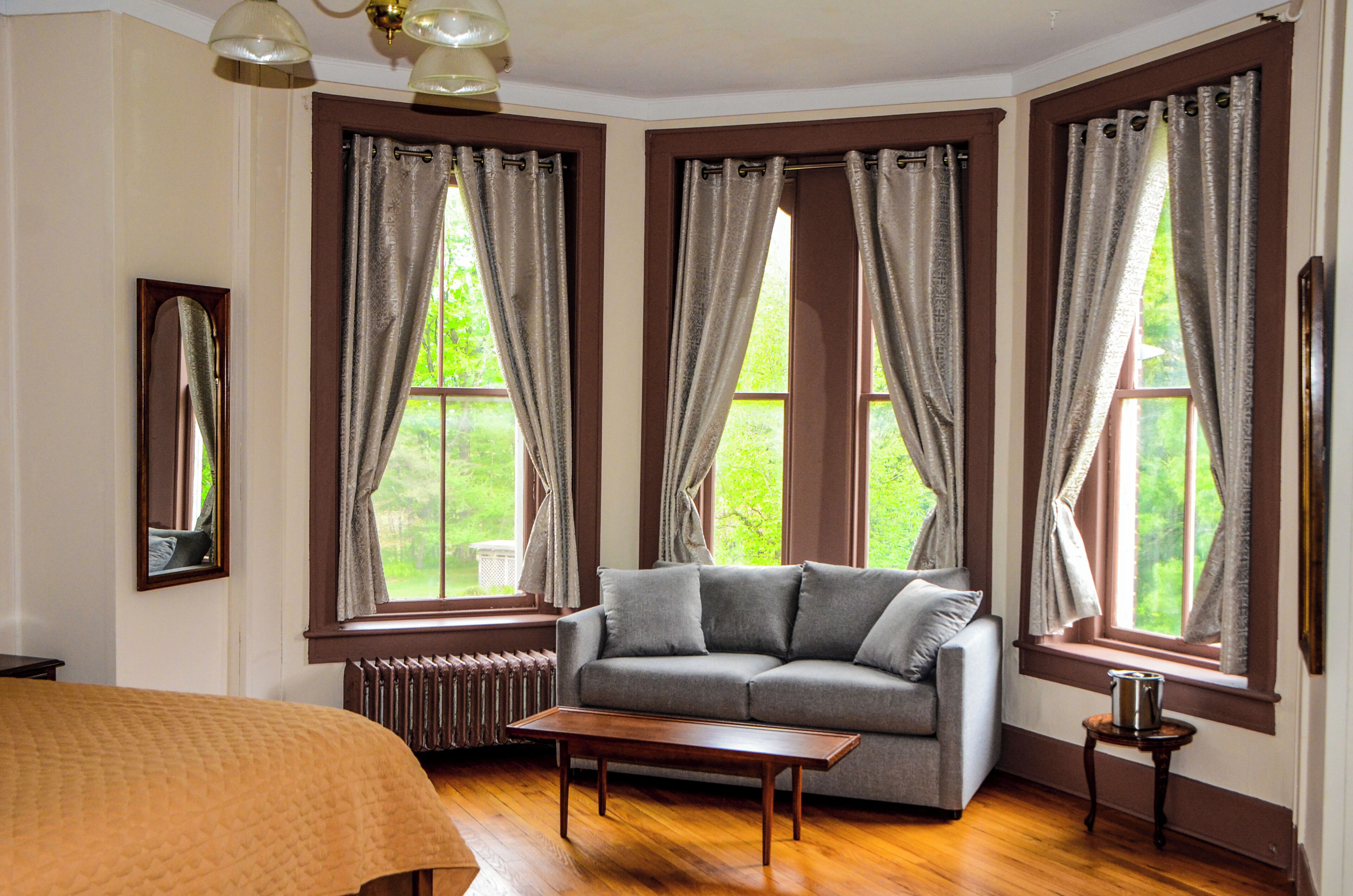Quarto com sofá cinzento, uma mesa de madeira e um radiador. Há três janelas com cortinas e um espelho na parede.