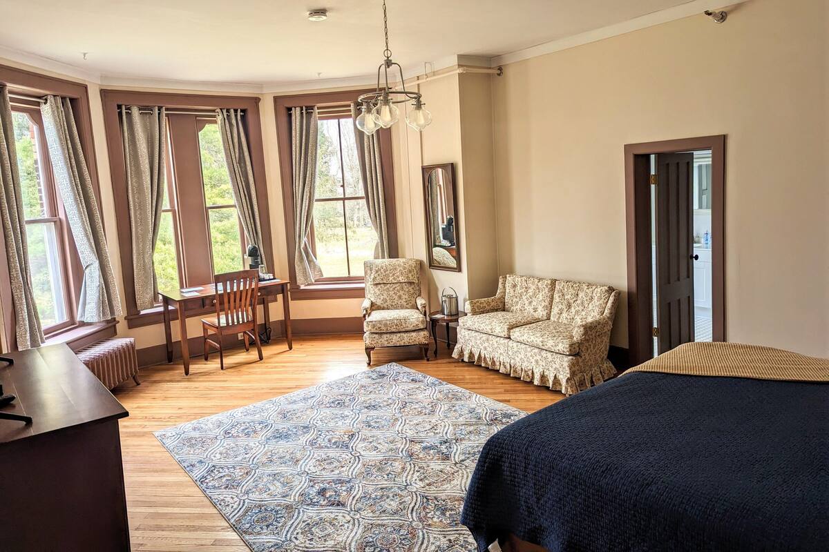Um quarto espaçoso com uma cama grande, um sofá, uma poltrona, uma escrivaninha e uma janela ampla com cortinas.