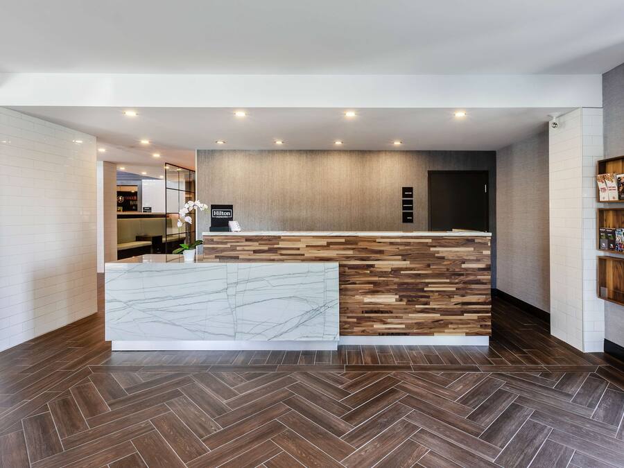 Uma área de recepção de hotel moderna, com piso de madeira em padrão herringbone, balcão de mármore e painel de parede de madeira.