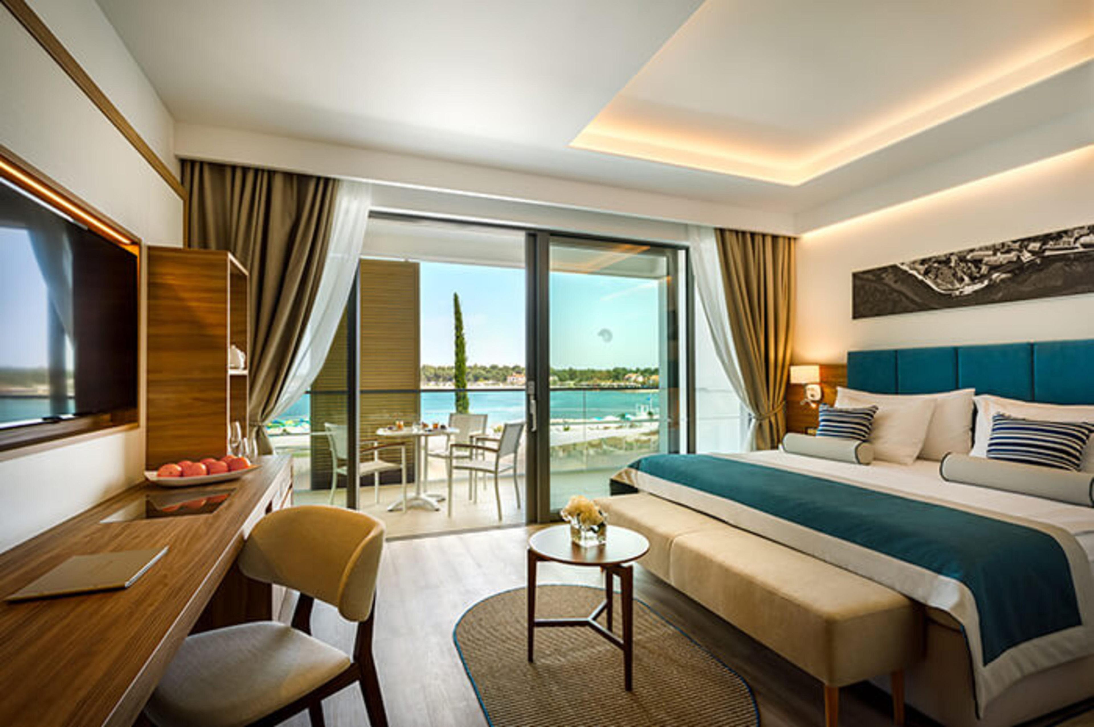Quarto de hotel com cama, escrivaninha, cadeira, mesa pequena e vista para o mar.