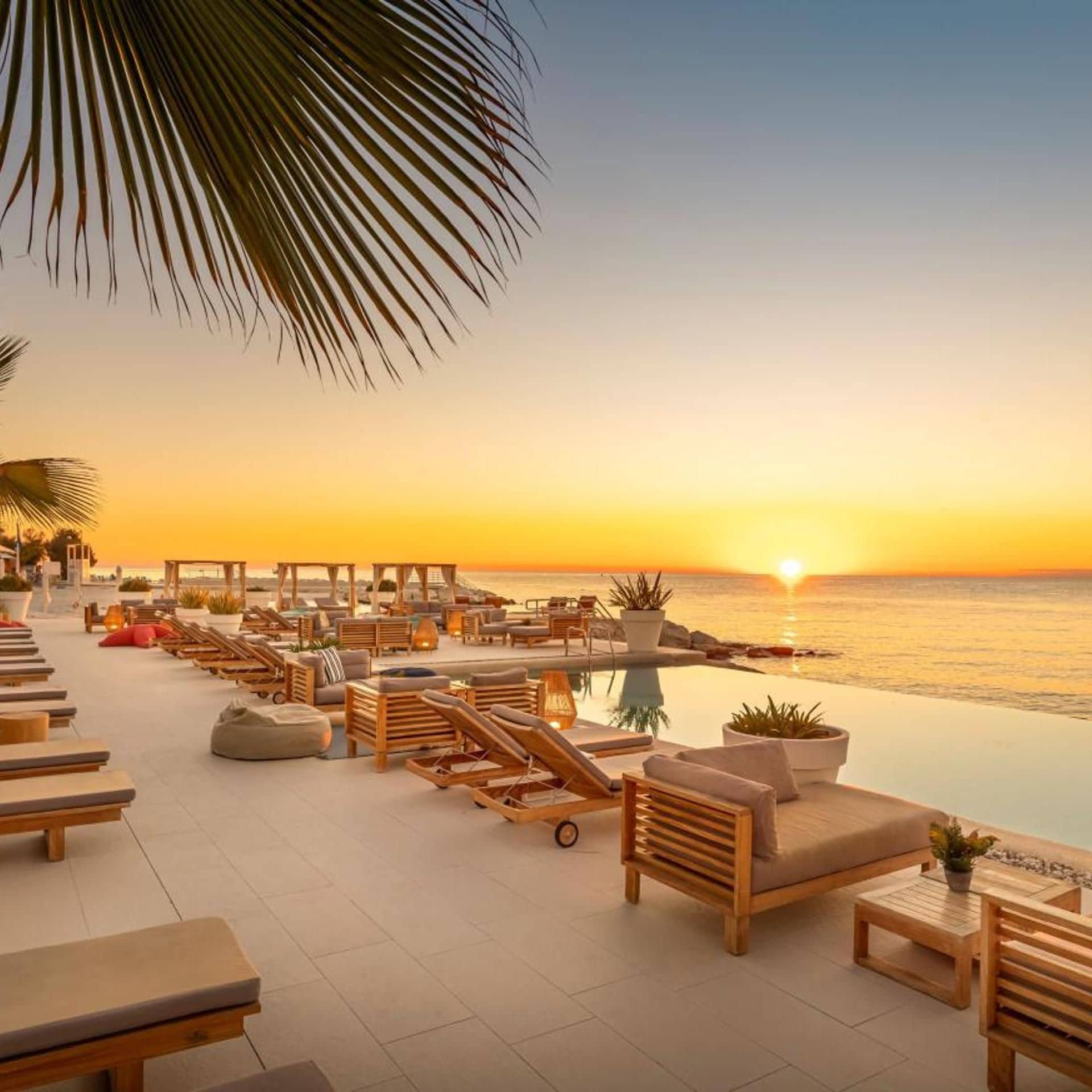 Uma área de estar à beira-mar com espreguiçadeiras, piscina e vista para o pôr do sol sobre o oceano.