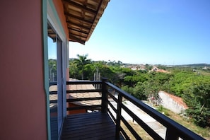 Uma varanda com piso e guarda-corpo de madeira, com vista para uma paisagem verdejante com árvores e edifícios ao longe.