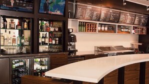 Uma área de bar com prateleiras cheias de garrafas de bebidas, uma televisão exibindo um canal de notícias e um quadro de cardápio.