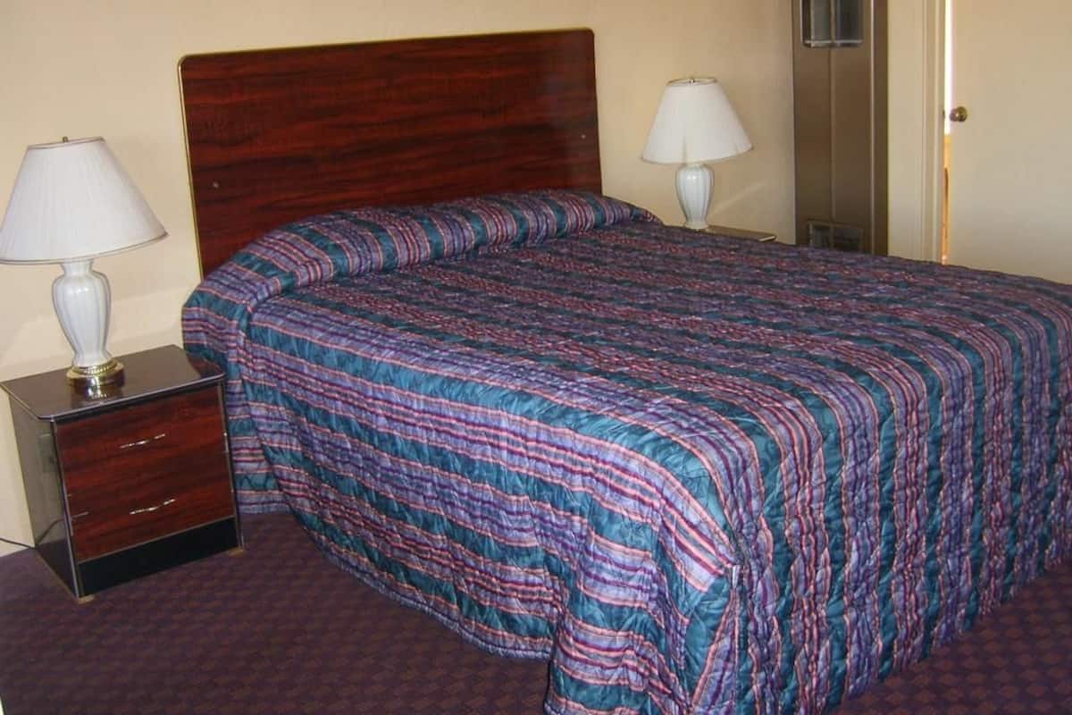 Uma cama bem arrumada com colcha listrada, cabeceira de madeira, duas luminárias de apoio e uma pequena mesa de cabeceira de madeira com gavetas.