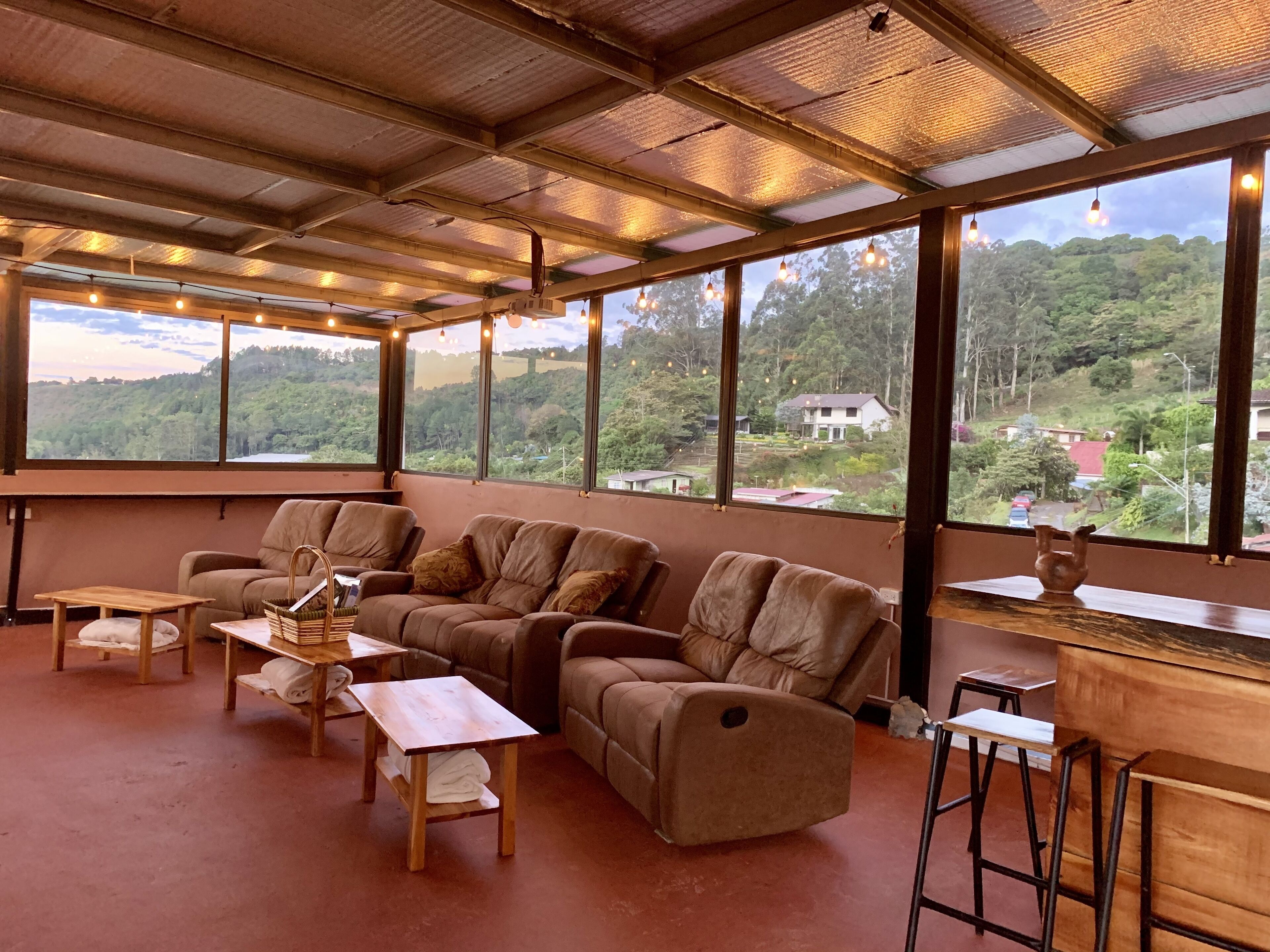 Uma área de estar acolhedora com sofá, mesas e bar. Grandes janelas oferecem vista para uma paisagem verde e exuberante.