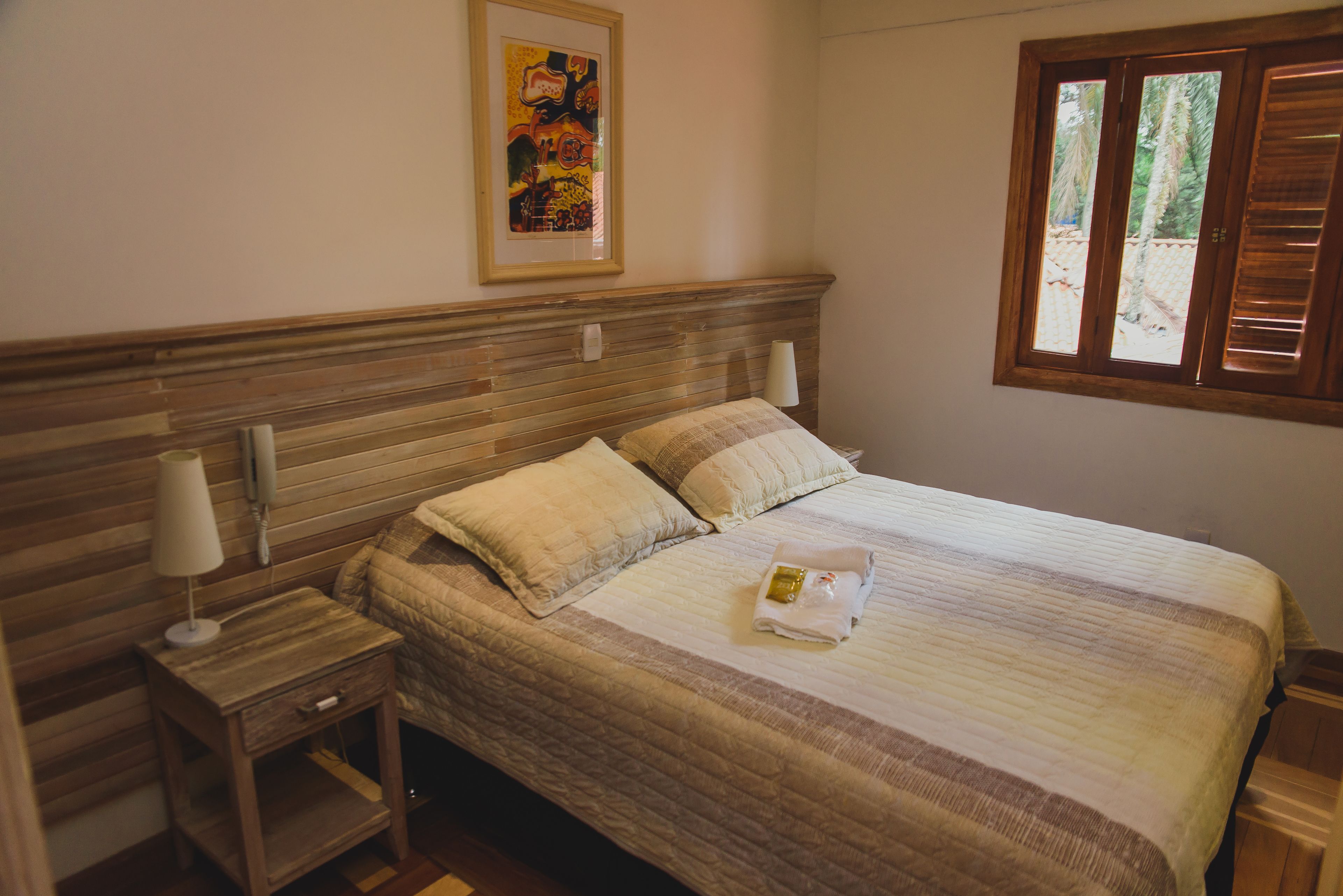 Cama bem arrumada com almofadas e uma toalha dobrada, mesa de cabeceira com abajur, e janela com persianas de madeira.
