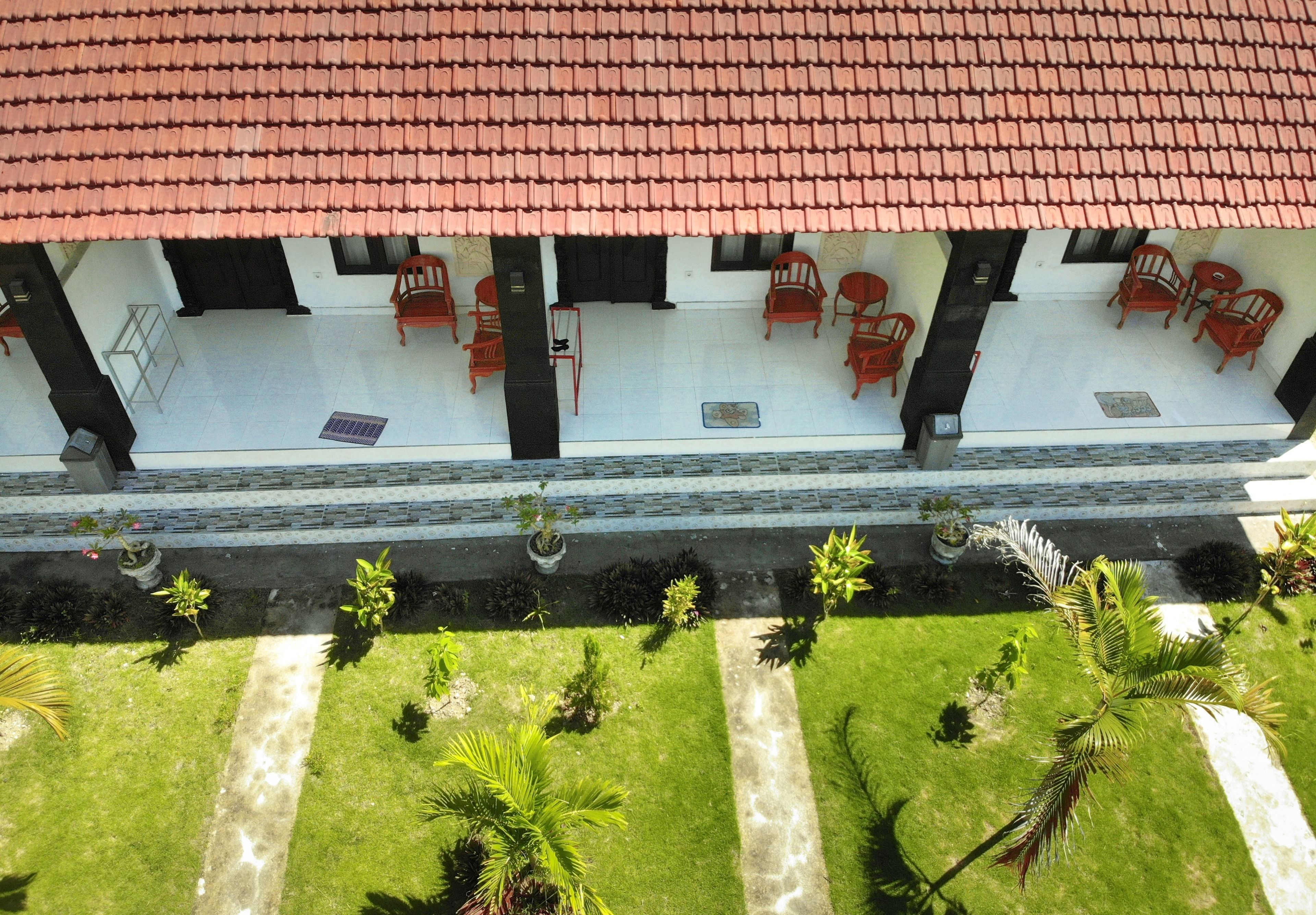 Um edifício com telhado de telhas, varanda coberta com cadeiras vermelhas e jardim com palmeiras.
